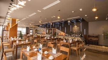 Restaurante moderno com mesas e cadeiras de madeira, área de bar e diversos vasos de plantas.