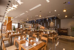 Restaurante moderno com mesas e cadeiras de madeira, área de bar e diversos vasos de plantas.
