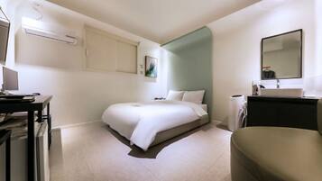 Quarto de hotel moderno com cama, escrivaninha com computador, mesa de cabeceira, espelho e banheiro com pia e vaso sanitário.
