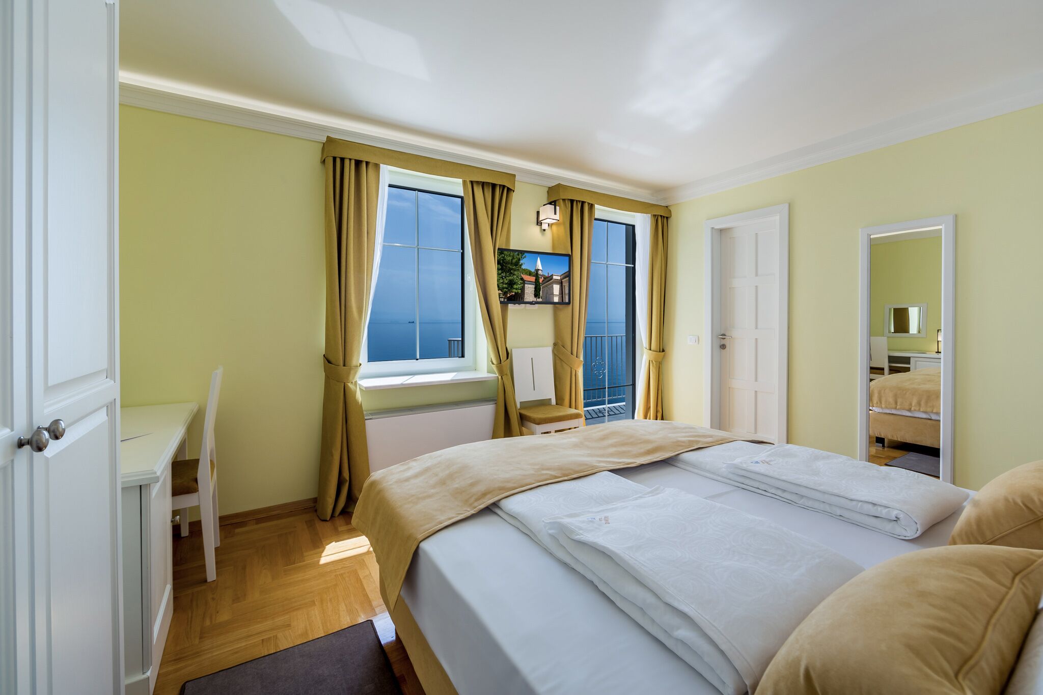 Quarto de hotel com cama, escrivaninha, cadeira, janela com cortinas e vista para o mar.