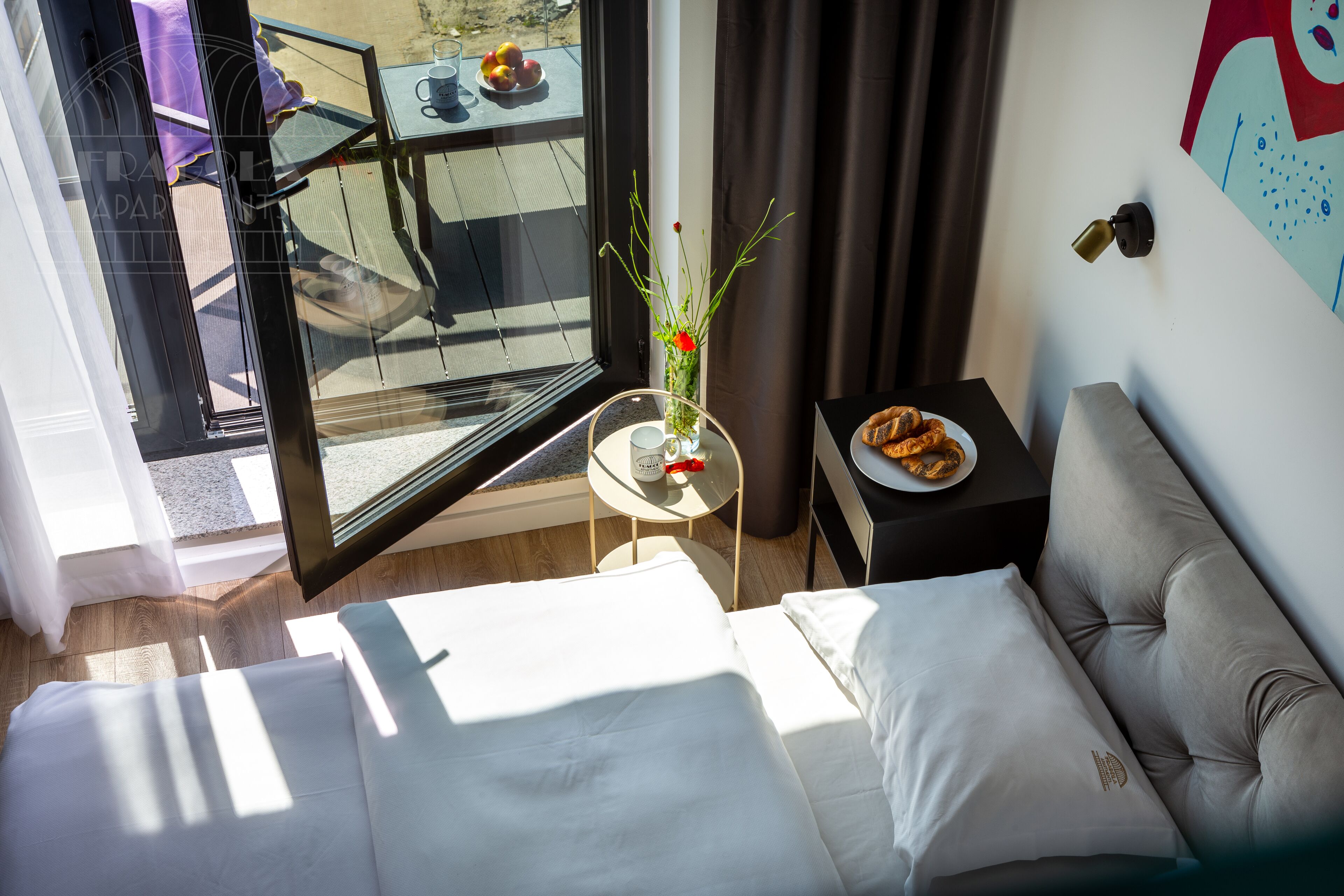 Quarto de hotel com cama, uma pequena mesa com um vaso, uma mesinha de cabeceira com um prato de comida e vista para a varanda.
