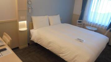 Quarto de hotel com uma cama grande, mesa de cabeceira, uma cadeira, uma mesinha redonda e uma janela com cortinas.