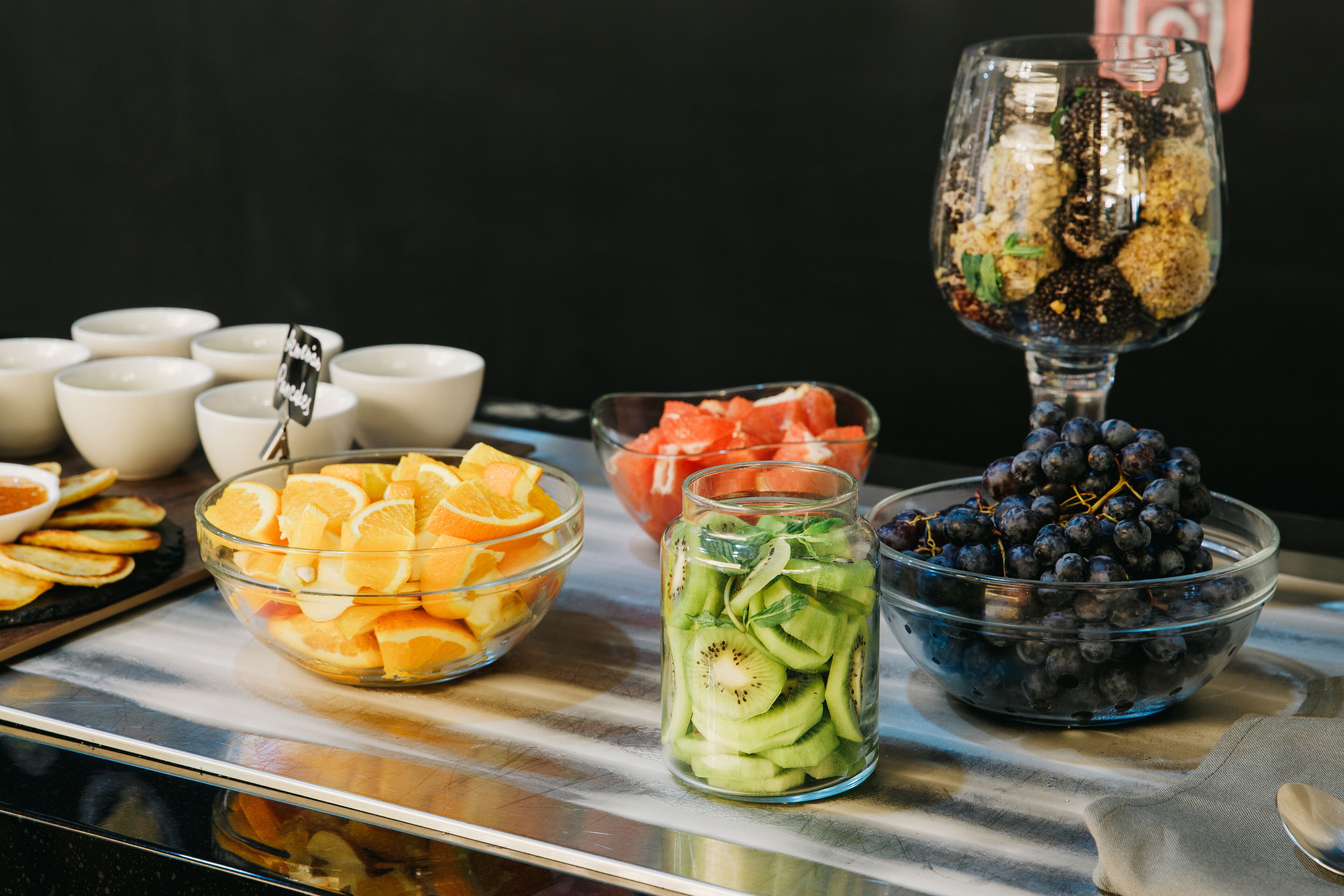 Uma mesa de buffet com diversos alimentos, incluindo tigelas com laranjas fatiadas, melancia, kiwi e uvas.