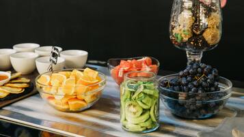 Uma mesa de buffet com diversos alimentos, incluindo tigelas com laranjas fatiadas, melancia, kiwi e uvas.