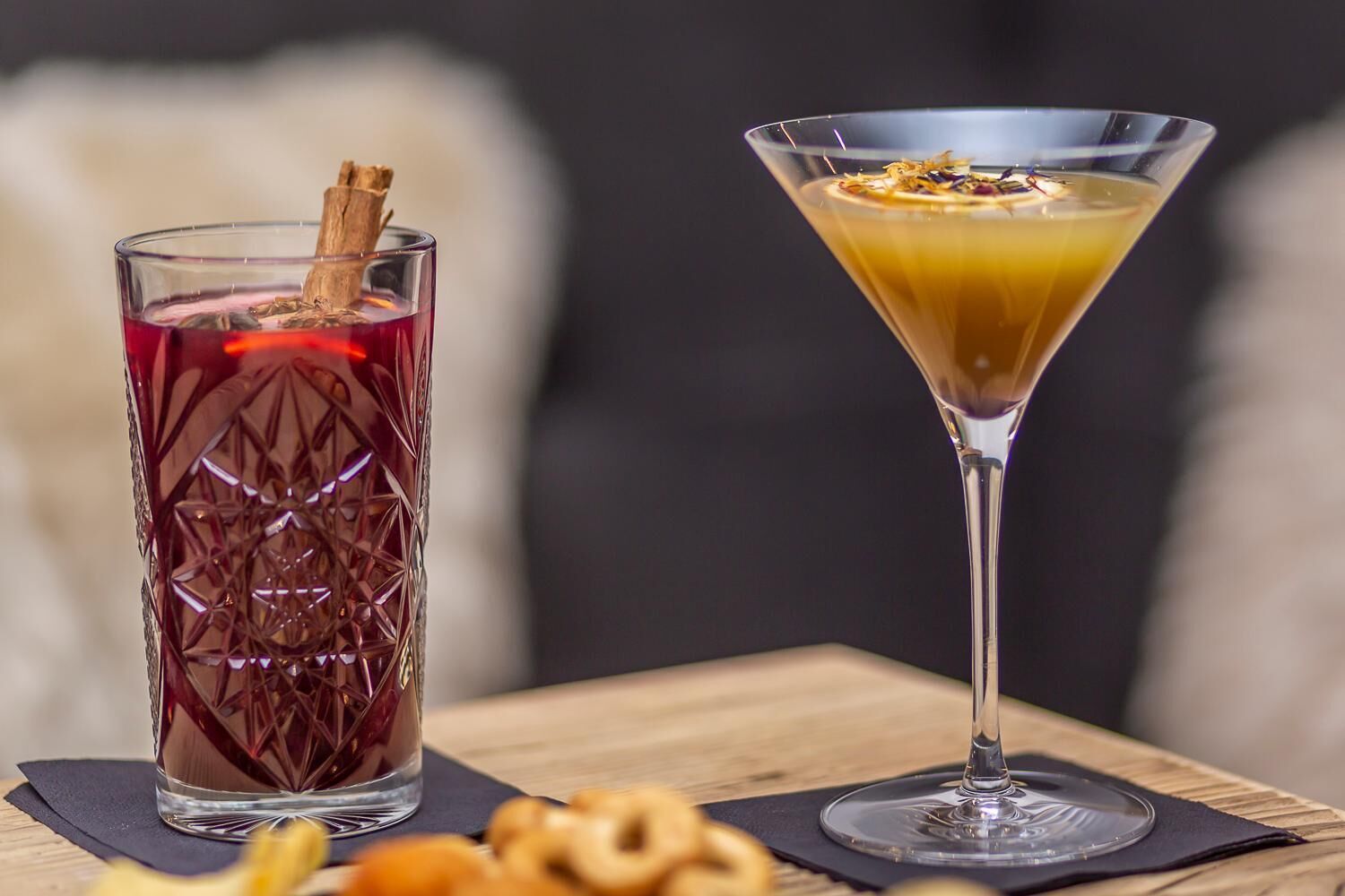 Duas bebidas em copos, uma com canela, sobre uma mesa de madeira com guardanapos e salgadinhos.