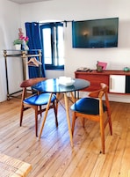 Uma sala de jantar com uma mesa redonda e quatro cadeiras, uma pequena mesa lateral com uma planta, uma TV fixada na parede e uma janela com cortinas azuis.