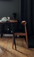 Piso de madeira, cortina escura, mesa com chapéu, planta e uma cadeira.