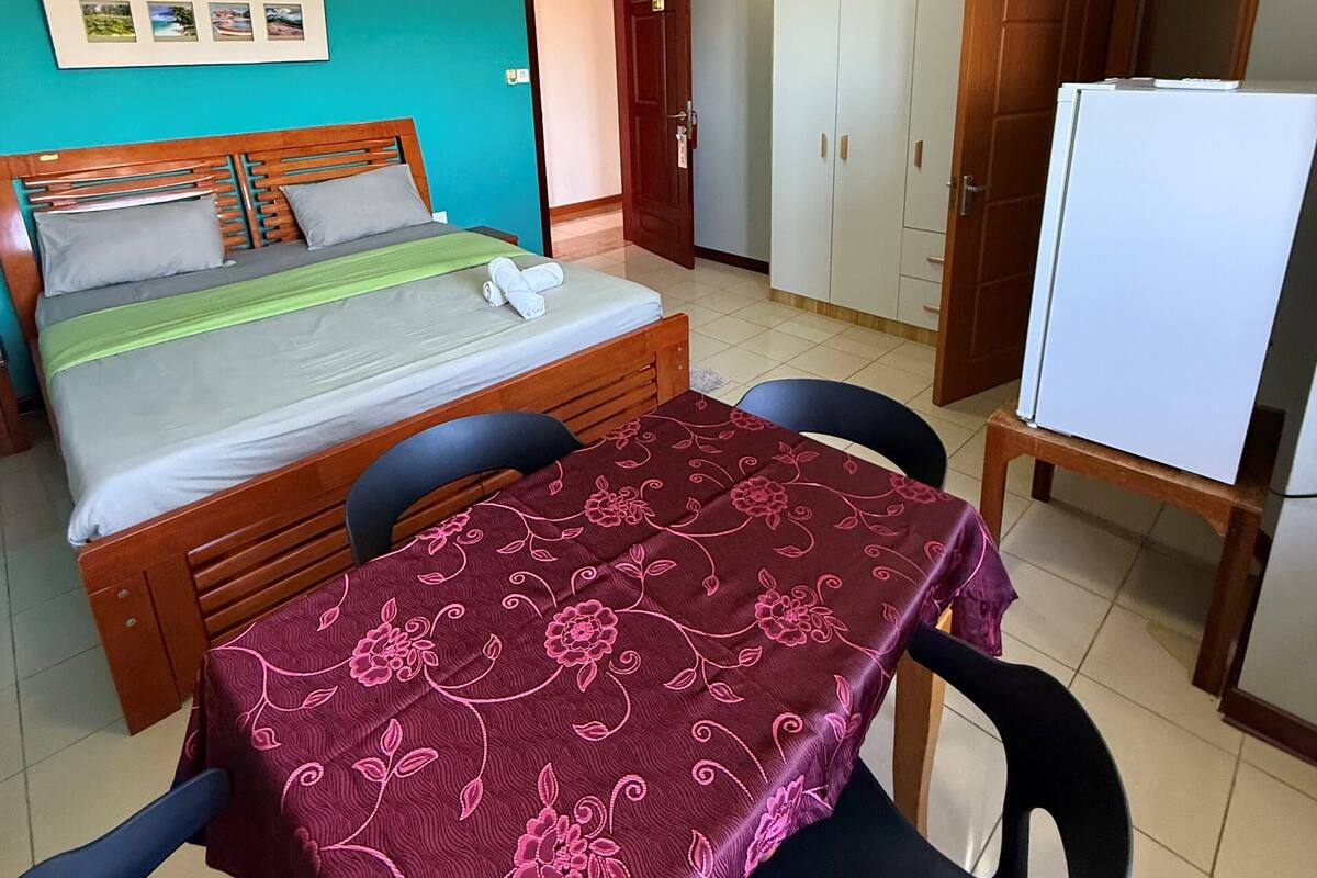 Um quarto com cama, mesa com cadeiras, guarda-roupa e frigorífico.