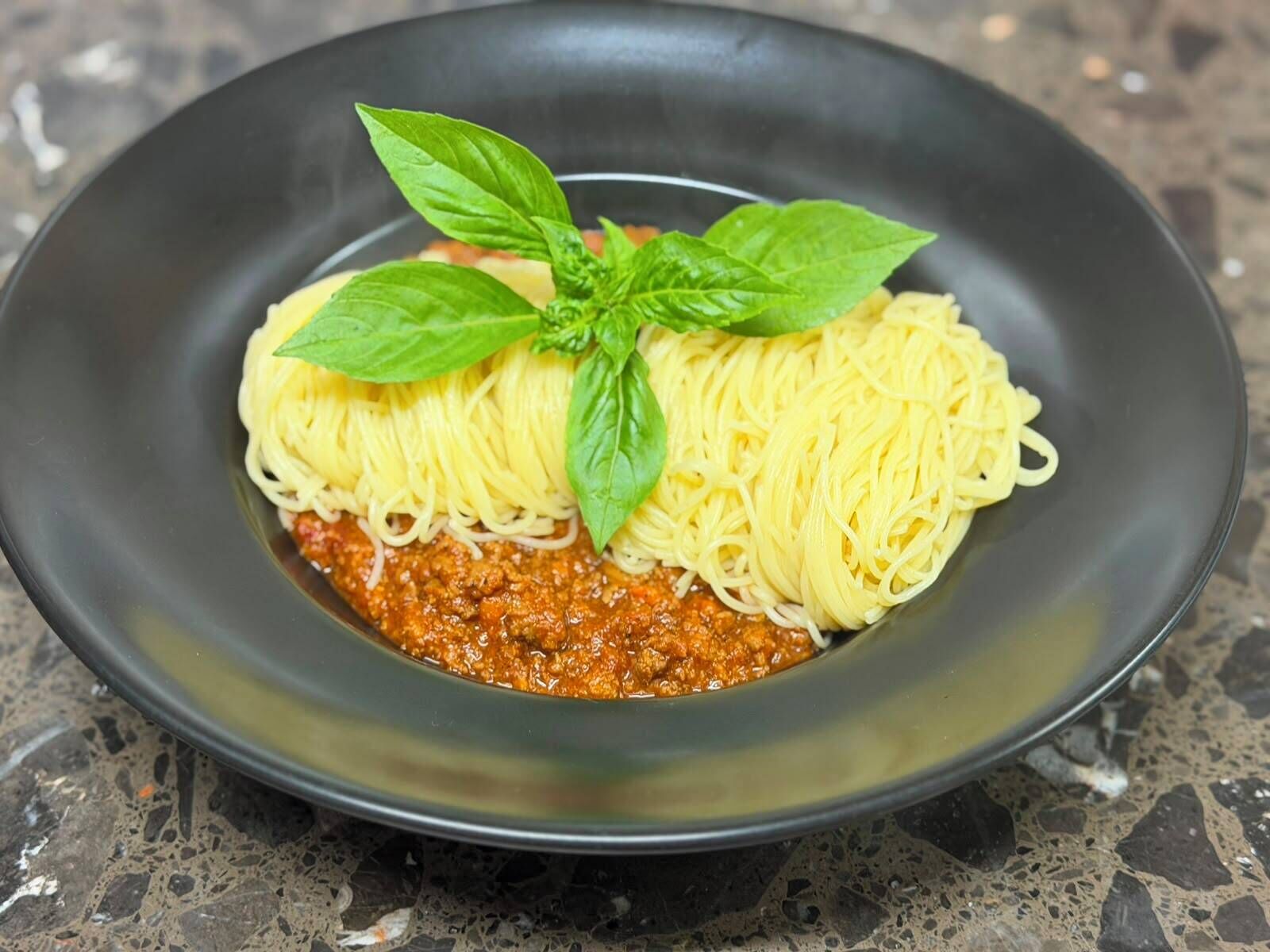 Uma tigela de espaguete com molho de tomate e folhas de manjericão sobre um prato escuro.