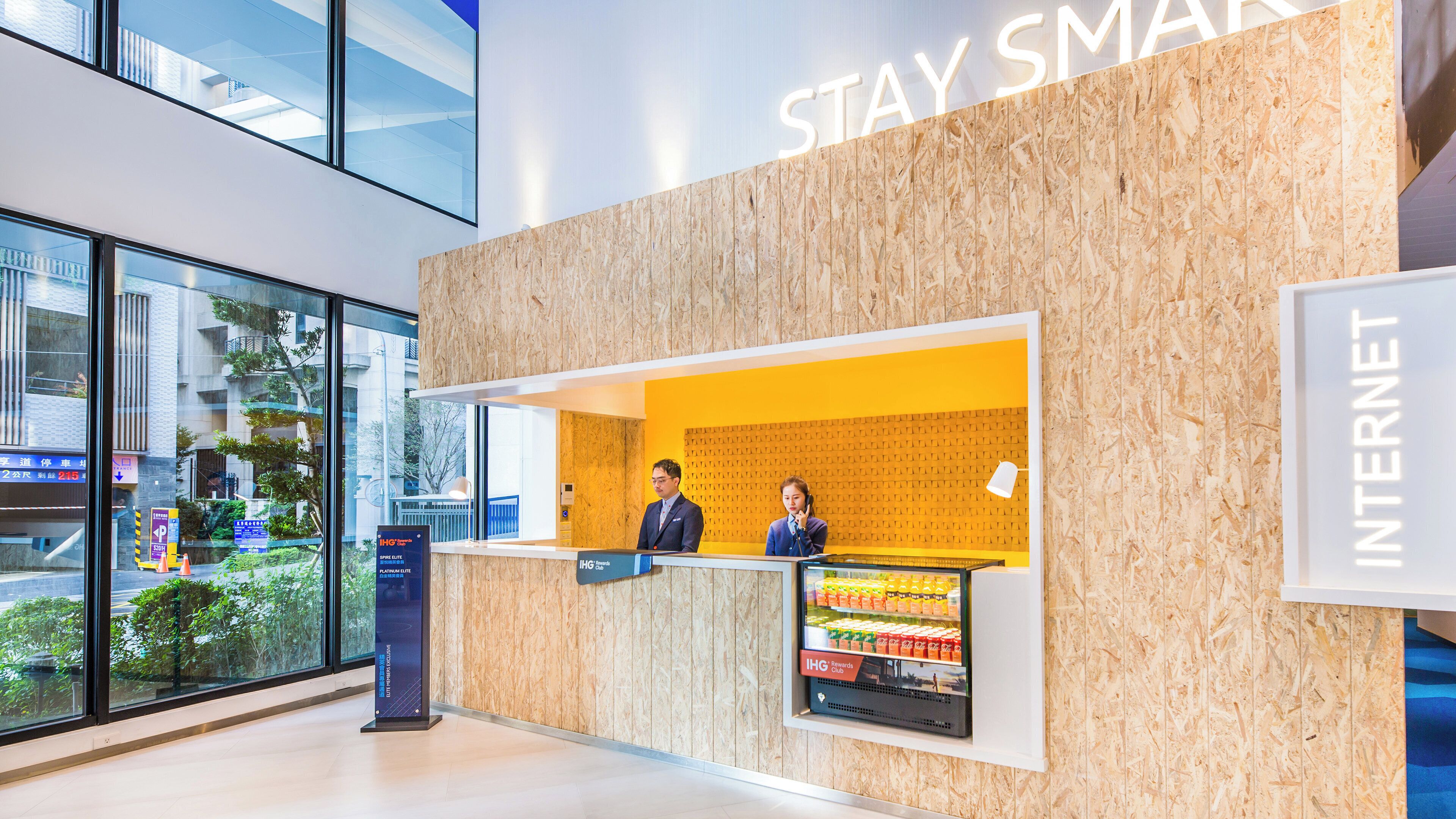 Balcão de recepção com design em painel de madeira, uma tela digital exibindo informações do hotel e vista para o exterior através de grandes janelas.