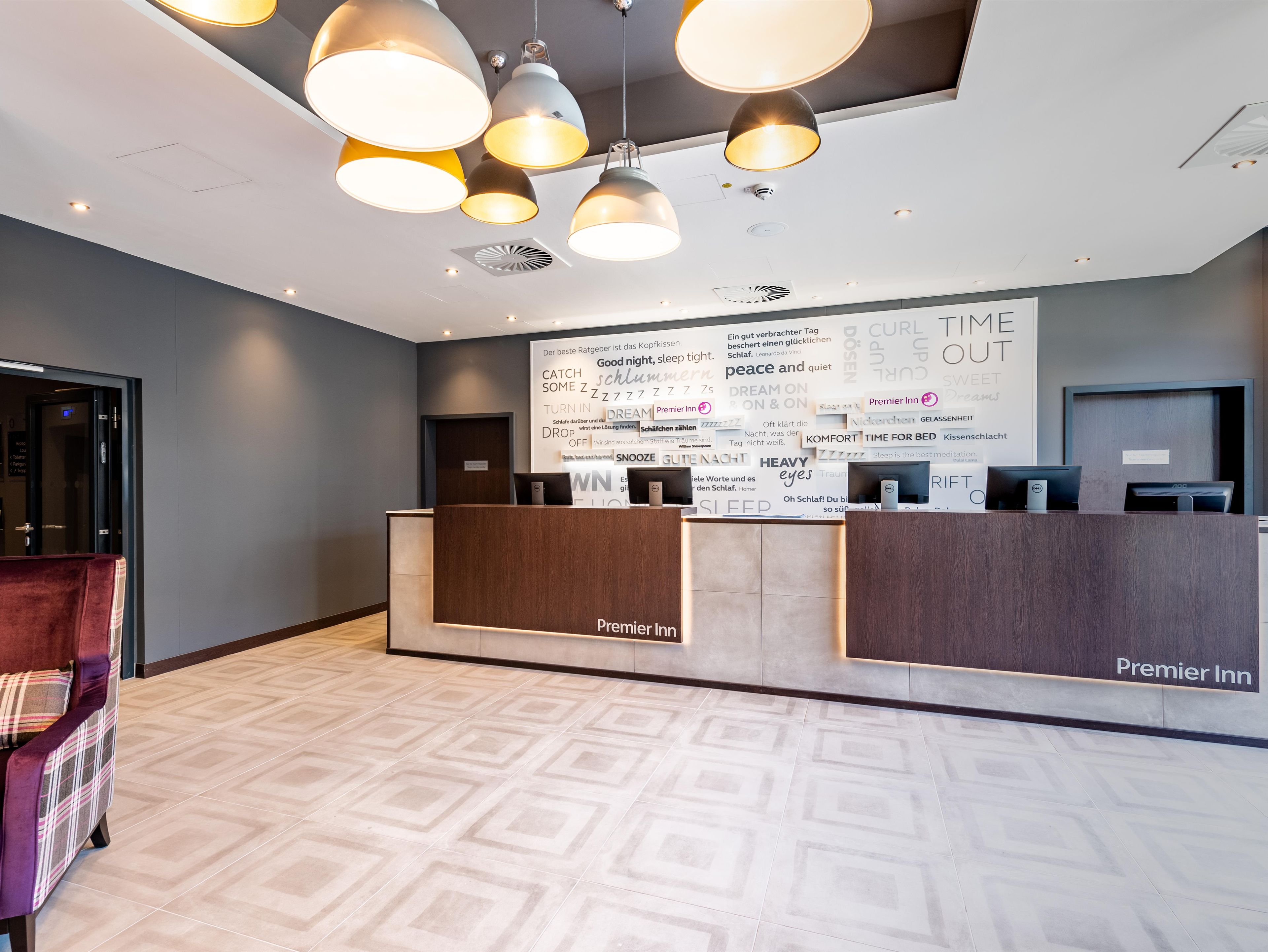 Uma área de recepção de hotel com design moderno, com balcão de atendimento, uma parede grande com frases motivacionais e piso estampado.