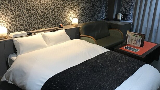 Quarto de hotel com cama, escrivaninha com luminária, uma cadeira e janela com persianas.