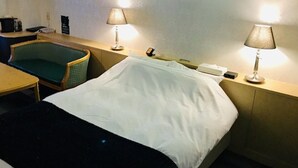 Quarto de hotel com cama, duas luminárias de cabeceira, uma cadeira, escrivaninha e televisão na parede.