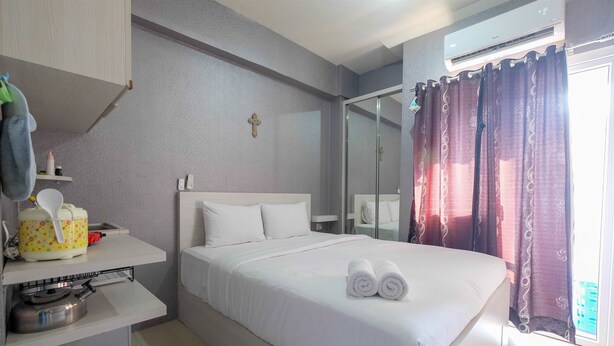 Quarto de hotel com uma cama, uma pequena mesa com chaleira, prateleira com produtos de higiene e uma janela com cortinas.