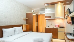 Quarto de hotel com uma cama, cabeceira de madeira, secretária embutida com espelho, pequeno kitchenette e banheiro com cortina de chuveiro.