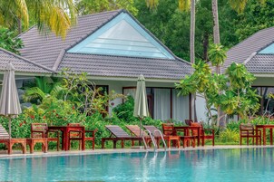 Área da piscina com espreguiçadeiras e mesas, cercada por vegetação exuberante e um edifício com telhado em azulejos.