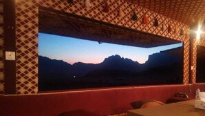 Quarto com uma janela grande que oferece vista para montanhas ao entardecer. Há uma mesa e um banco com almofada estampada.