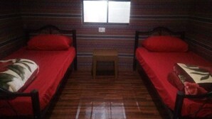 Quarto com duas camas de solteiro, cada uma com roupa de cama vermelha e uma almofada. Há uma pequena mesa de madeira no centro.