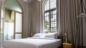 Quarto de hotel com uma cama grande, um ventilador de teto, uma janela com cortinas e uma pequena mesa de cabeceira de madeira com uma garrafa e uma planta.
