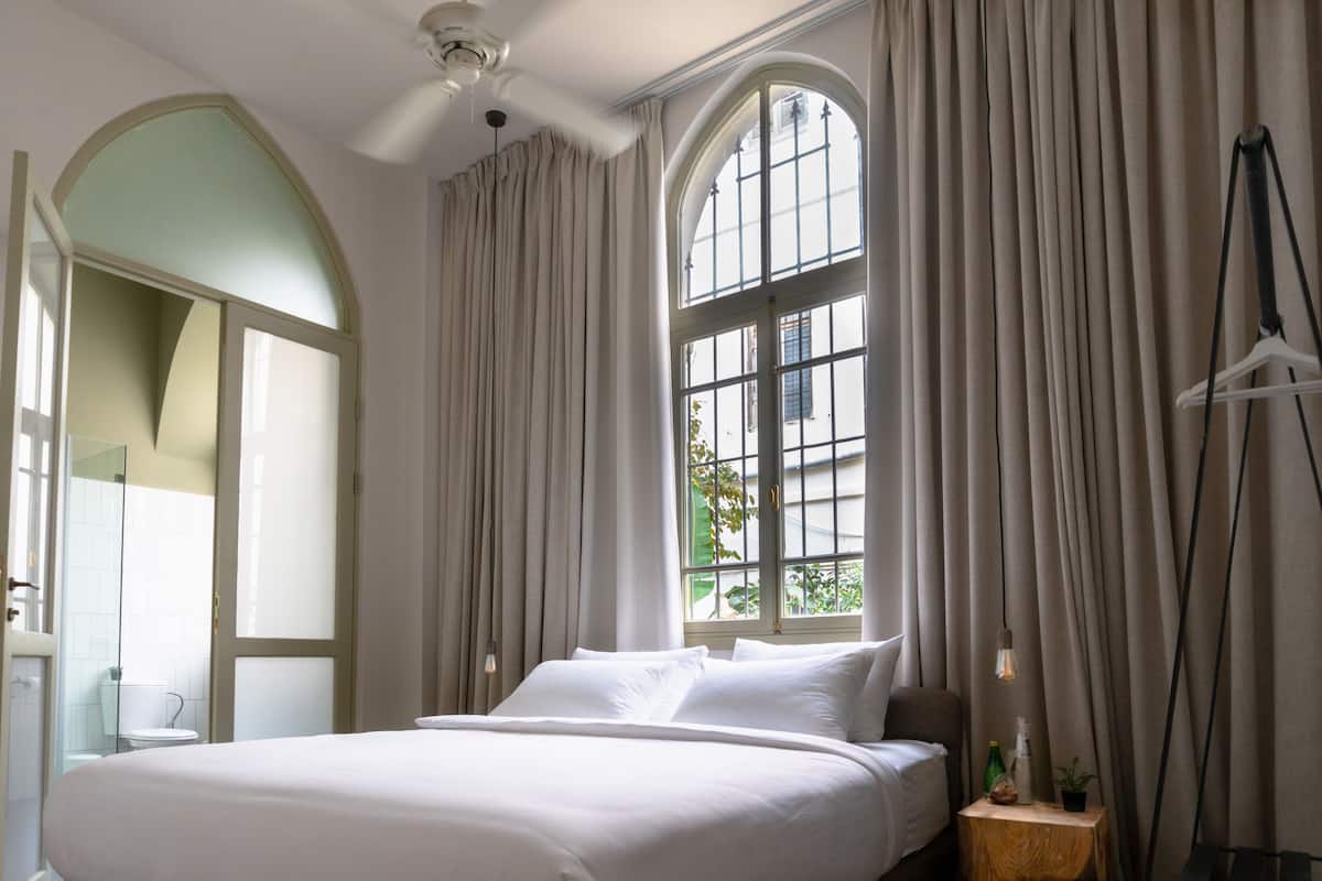 Quarto de hotel com uma cama grande, um ventilador de teto, uma janela com cortinas e uma pequena mesa de cabeceira de madeira com uma garrafa e uma planta.