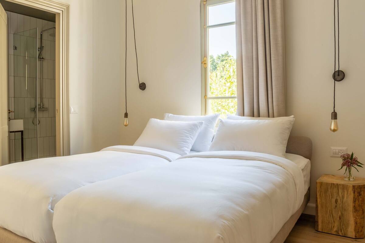 Uma cama grande com roupa de cama branca, uma mesa de cabeceira de madeira com um vaso e uma janela com cortinas.