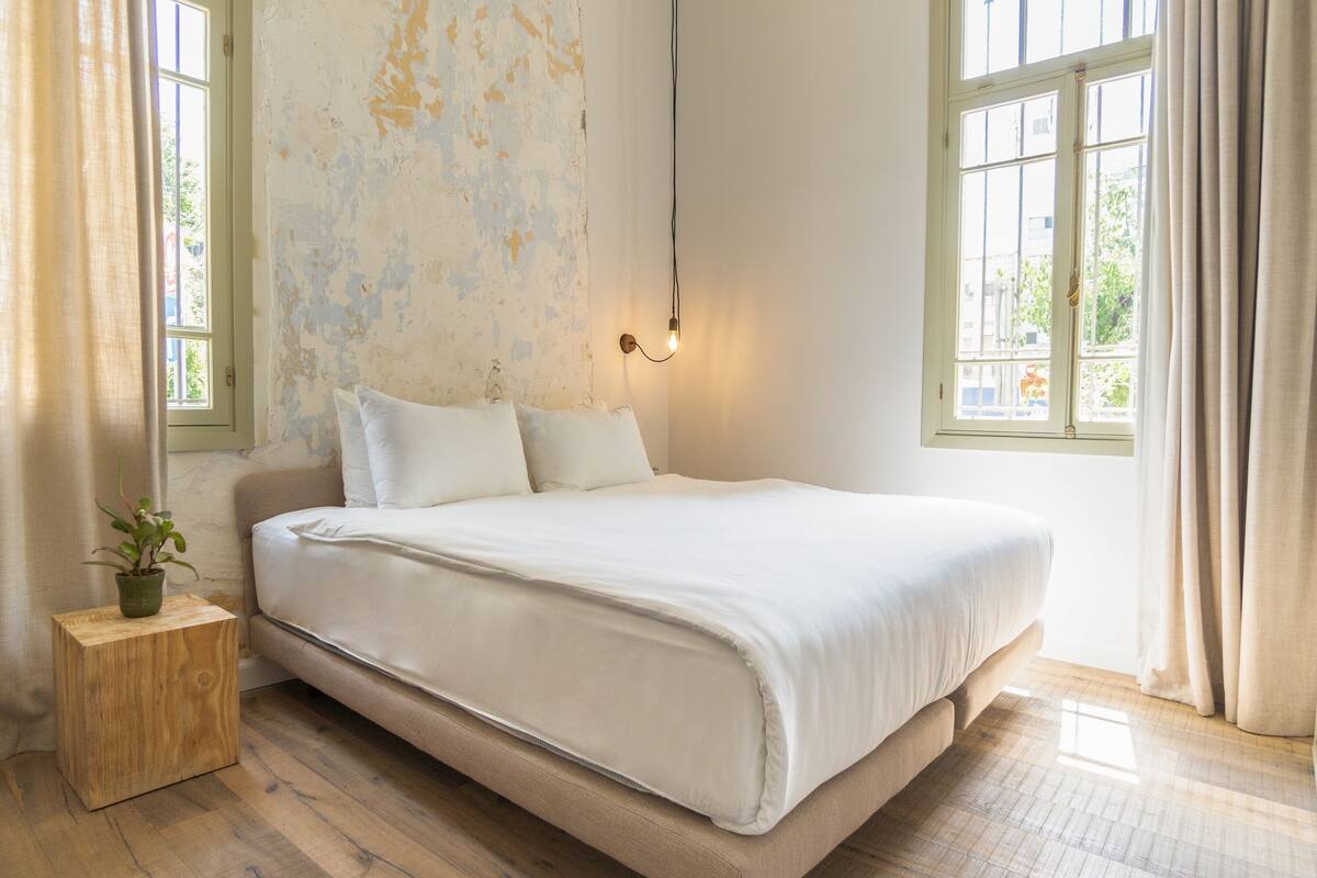 Quarto com uma cama, uma pequena mesa de cabeceira de madeira com uma planta, e uma parede com tinta descascada.