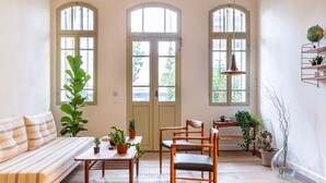 Uma sala de estar aconchegante com um sofá, uma mesa de madeira e cadeiras. Há janelas grandes com arcos, um abajur pendente e vários vasos de plantas.