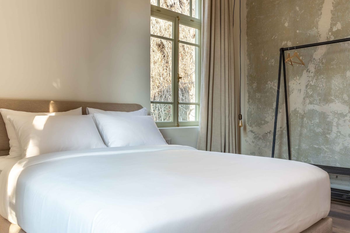 Cama com roupa de cama branca, janela com cortinas e um suporte de metal encostado em uma parede com textura.