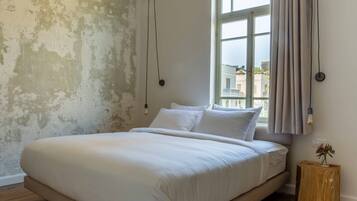 Quarto com uma cama grande, mesa de cabeceira de madeira, janela com cortinas e parede com textura desgastada.