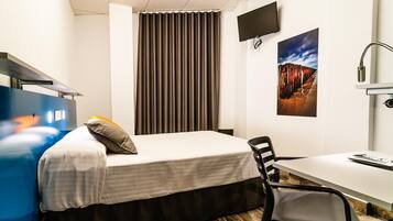 Quarto de hotel com cama, escrivaninha, cadeira, televisão e janela com cortinas.