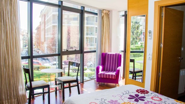 Quarto de hotel com grandes janelas, cama com colcha florida, cadeira roxa e uma pequena mesa.