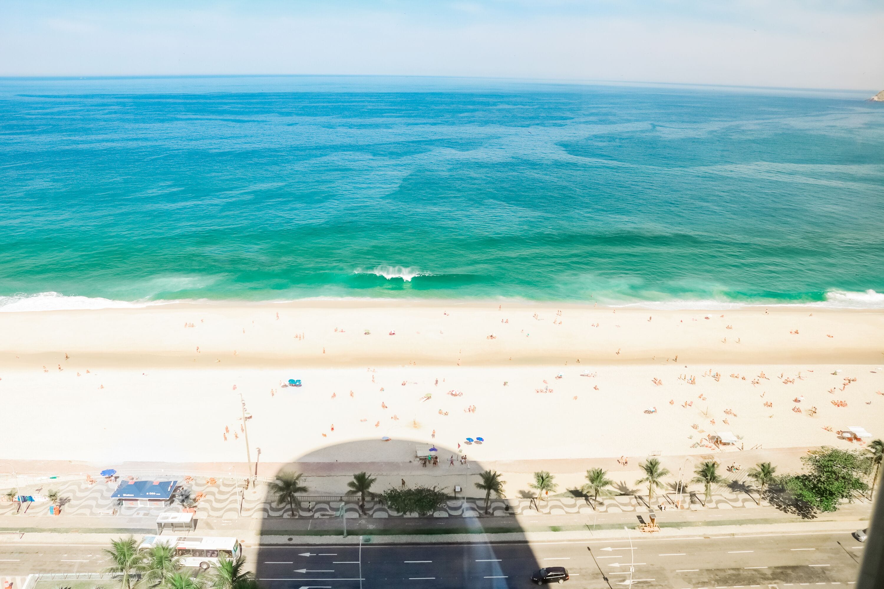 Vista aérea de uma praia com uma estrada em primeiro plano, palmeiras e uma visão clara do oceano.