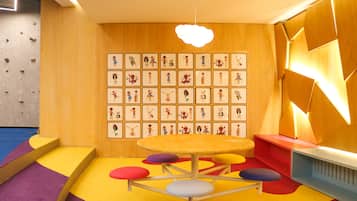 Uma sala de jogos com uma parede de escalada, tapetes de jogo coloridos, uma mesa redonda com banquetas e uma parede com ilustrações emolduradas.
