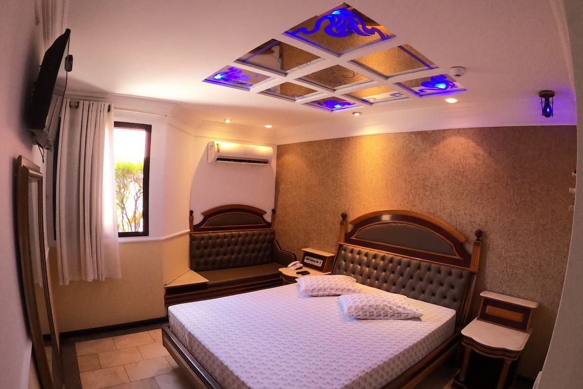 Quarto de hotel com uma cama grande, mesa de cabeceira, espelho, TV fixada na parede e teto com iluminação decorativa.