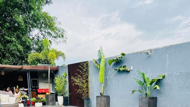 Uma área à beira da piscina com mobiliário de vime, incluindo comida e bebidas, cercada por plantas em vasos e um edifício ao fundo.