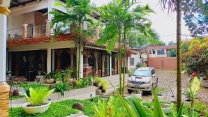 Uma casa de dois andares com varanda coberta, cercada por vegetação exuberante e plantas em vasos.