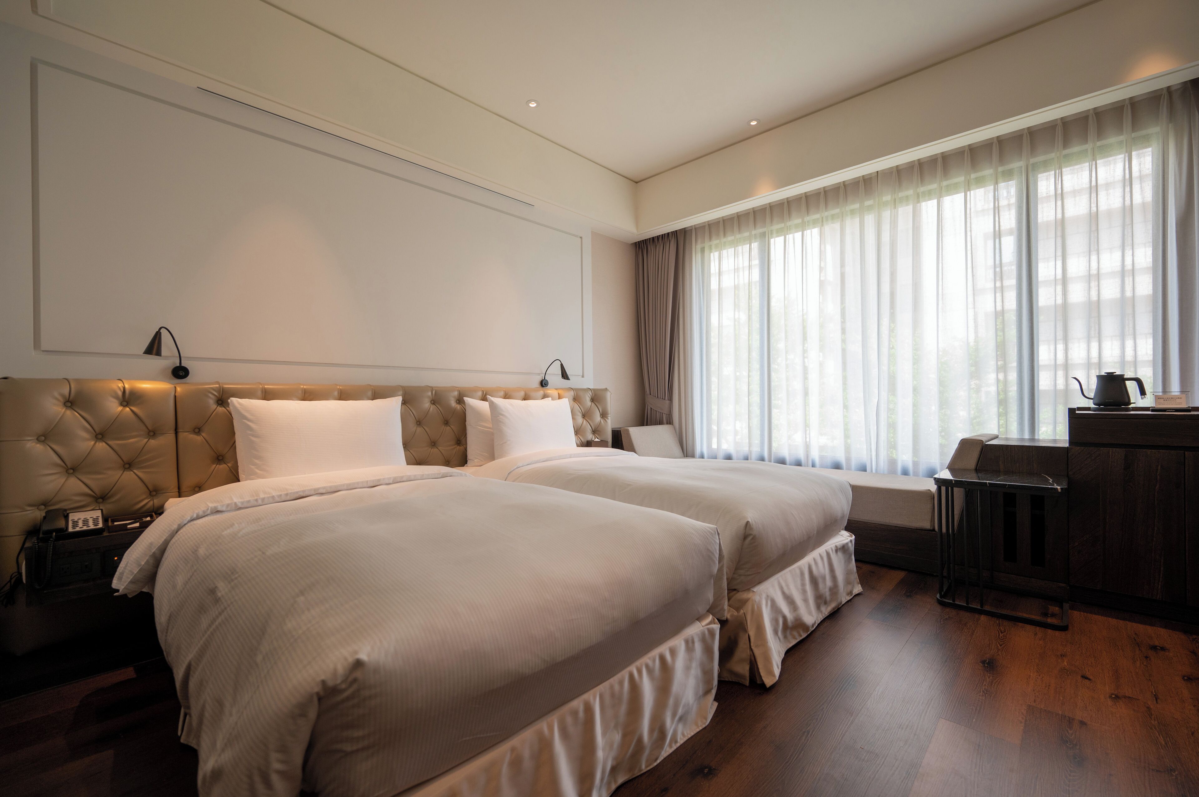 Quarto de hotel com duas camas, uma janela grande com cortinas, mesa de cabeceira com abajur e um pequeno armário.