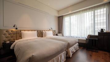 Quarto de hotel com duas camas, uma janela grande com cortinas, mesa de cabeceira com abajur e um pequeno armário.