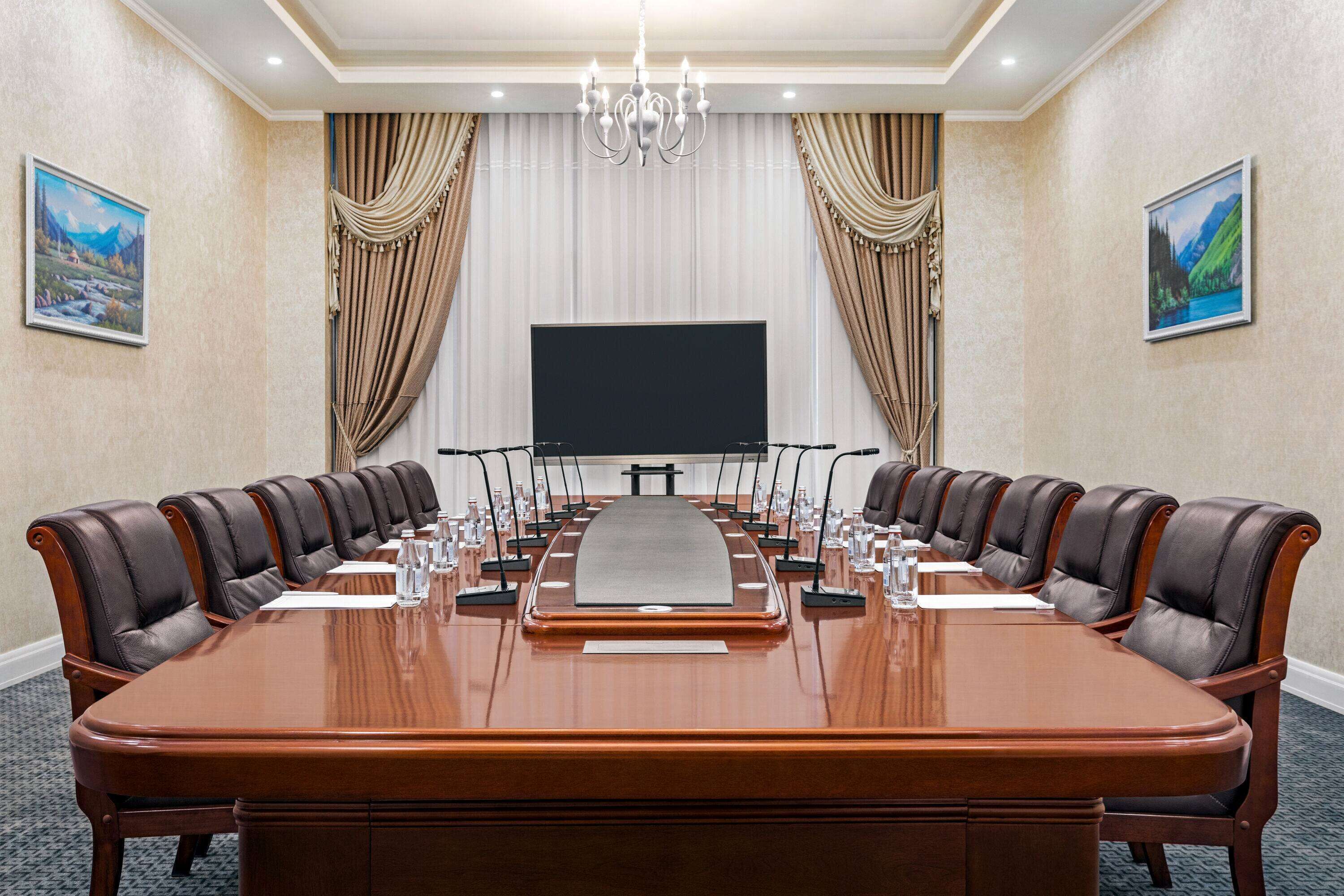 Uma sala de conferências com uma mesa comprida, cadeiras de couro, microfones e uma tela grande.