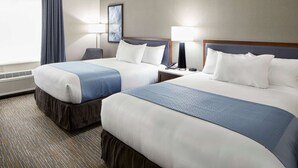 Quarto de hotel com duas camas, uma escrivaninha, uma cadeira, uma luminária e uma janela com cortinas.