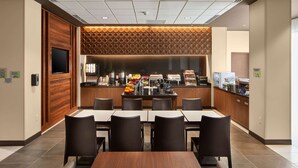 Quarto de hotel moderno com área de refeições, mesa e cadeiras de madeira, um espaço para café da manhã com bebidas e lanches diversos, e uma grande janela que oferece luz natural.