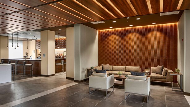 Um lobby moderno com teto de madeira, área de estar com sofás e cadeiras, e um bar com banquetas.