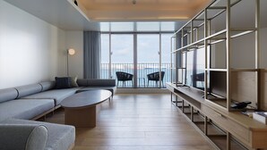 Uma sala de estar moderna com sofá, mesa de centro e aparador de TV, grandes janelas com vista para o mar, e uma varanda com cadeiras.