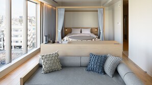 Quarto de hotel moderno com uma janela grande, cama com cabeceira de madeira, sofá com almofadas e vista para a cidade.