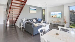 Uma sala de estar moderna com um sofá cinzento, uma mesa de centro e uma televisão. Há uma escada de madeira com corrimão.