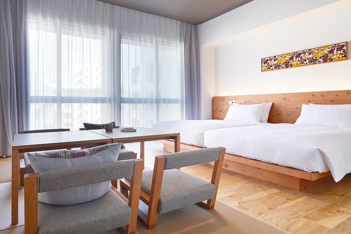 Quarto de hotel com cama de madeira, uma mesa pequena, duas cadeiras, uma janela grande com cortinas leves e uma obra de arte colorida na parede.