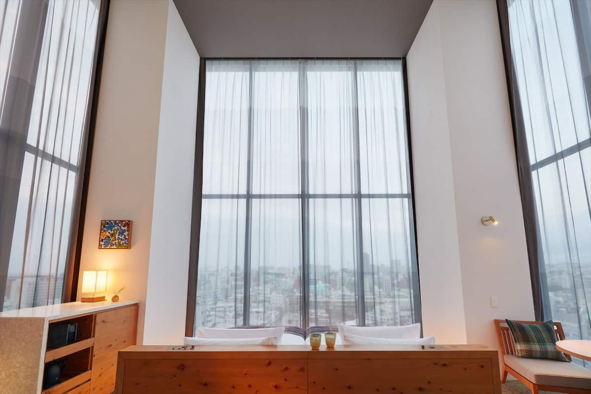 Quarto de hotel moderno com uma janela grande, uma cama de madeira, uma mesinha de cabeceira com abajur e vista para a paisagem urbana.