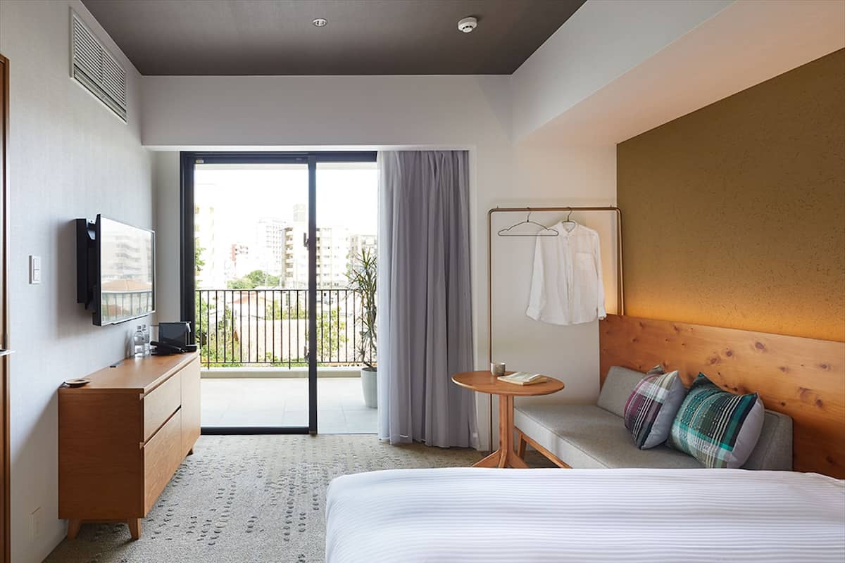 Quarto de hotel com cama, escrivaninha, cadeira, televisão, varanda com vista para edifícios e um cabideiro.