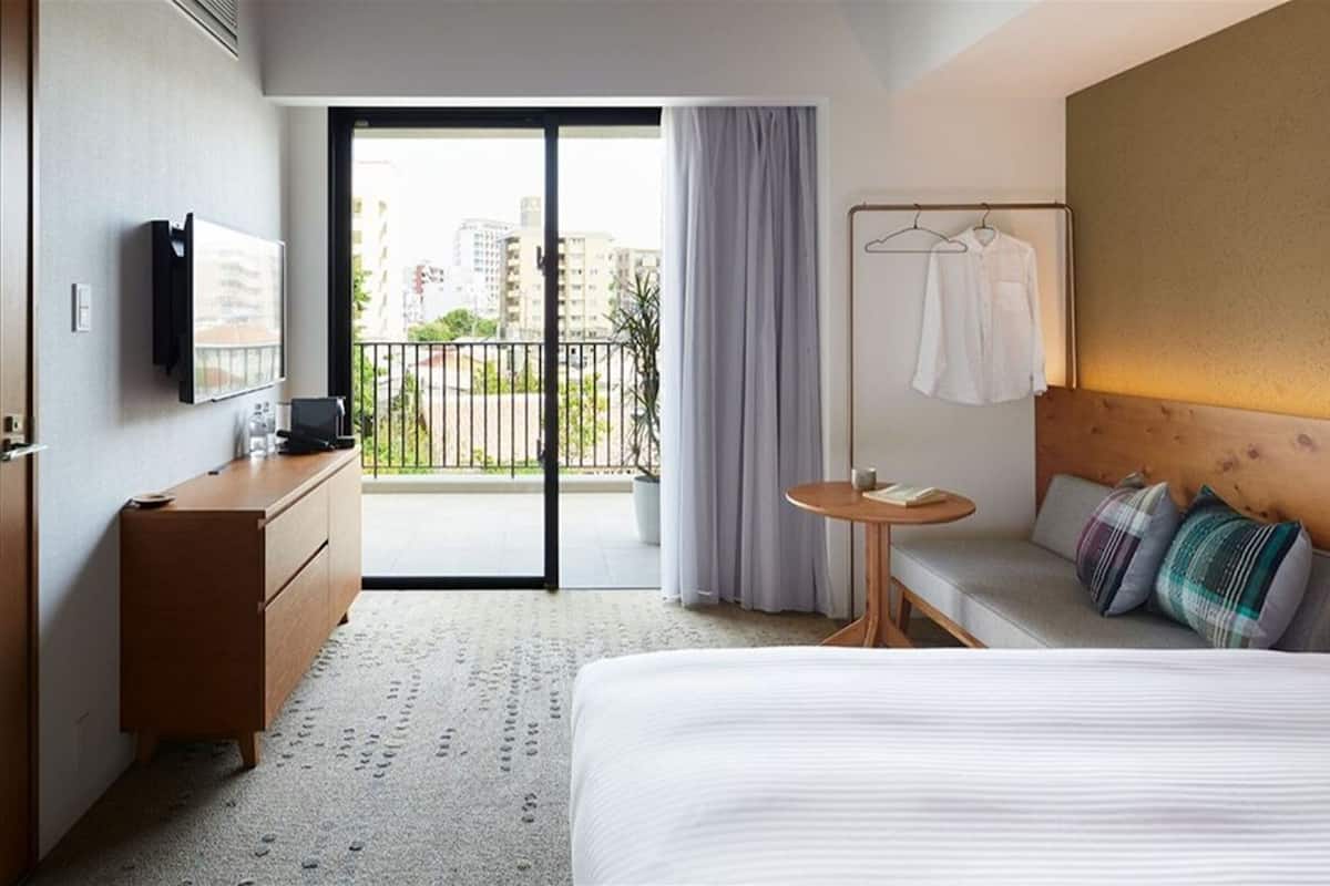 Quarto de hotel com cama, escrivaninha, cadeira, mesa pequena, televisão, varanda com vista para edifícios e um cabideiro.