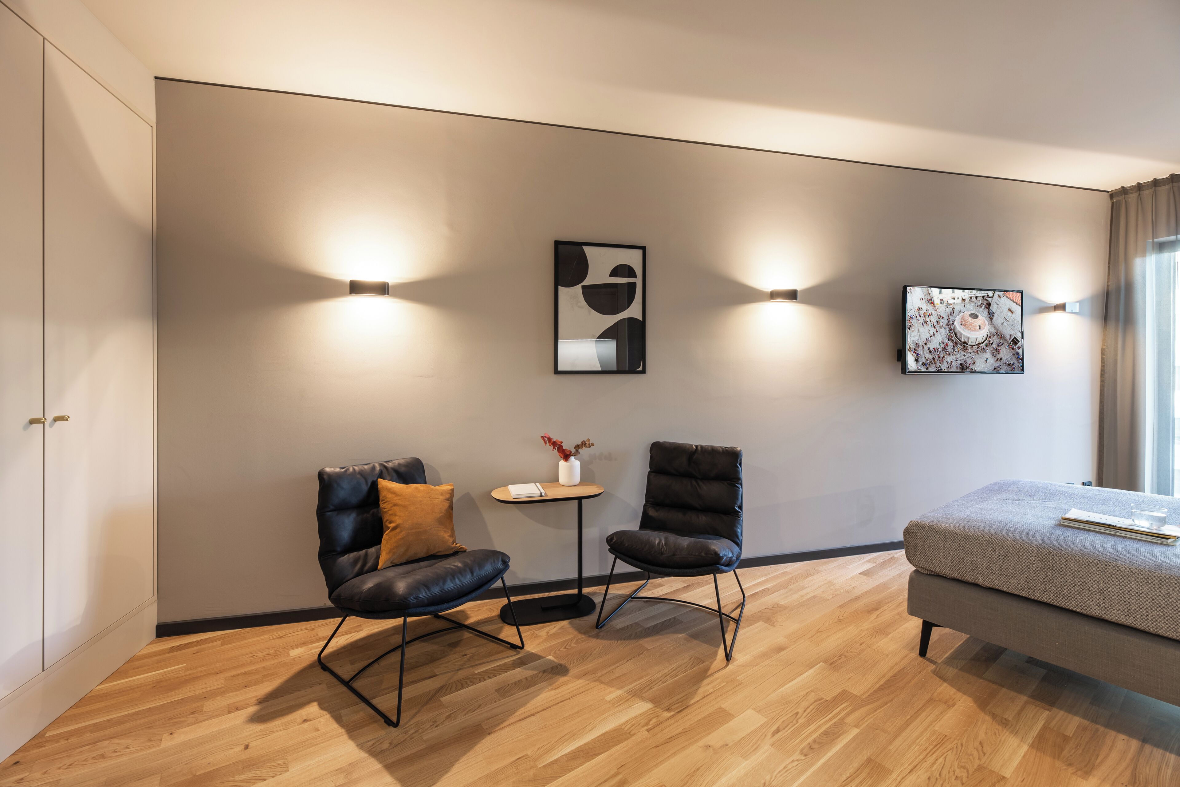 Uma sala de estar moderna com duas poltronas de couro, uma mesa redonda pequena e um sofá. Há duas luminárias fixadas na parede e duas obras de arte emolduradas.